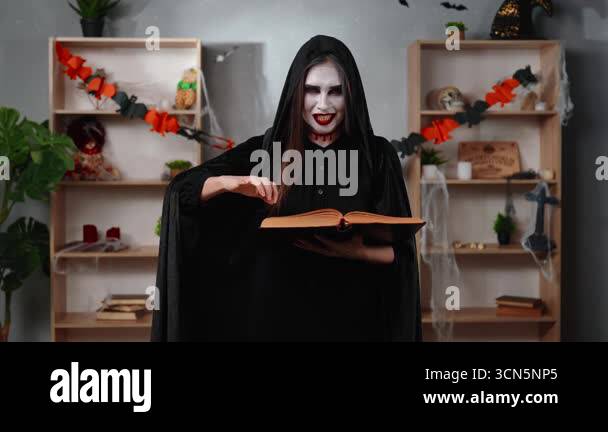 A woman with vampire makeup and a witch hat reads spells from a magic ...