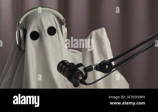 A person dressed as a ghost, wearing a white sheet and headphones, sits ...