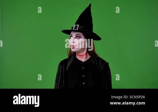 A witch with vampire makeup, in black cloak and hat, points her fingers ...