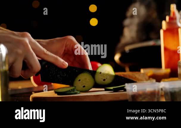 A chef skillfully slices a fresh cucumber on a wooden board in a dimly ...