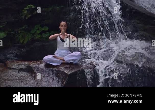 Young woman sitting in lotus pose under tropical waterfall meditating ...