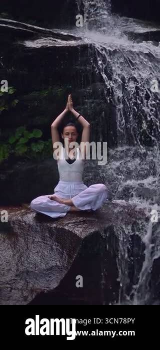 Woman sitting in lotus pose under tropical waterfall meditating with ...