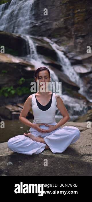 Woman sitting in lotus pose under tropical waterfall meditating with ...