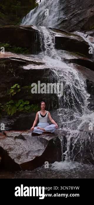 Woman sitting in lotus pose under tropical waterfall meditating with ...