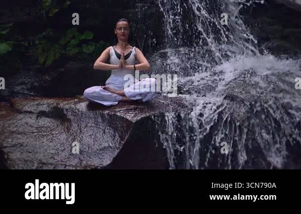 Young woman sitting in lotus pose under tropical waterfall meditating ...