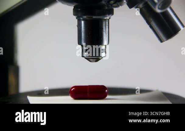 A microscope lens is lowered to a red pill and a male doctor's gloved ...