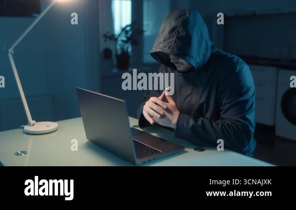 Portrait of a mysterious male hacker working on his laptop in a dimly lit room. The dark ...