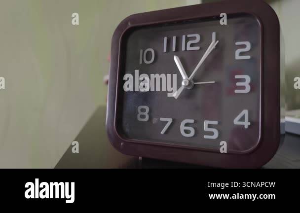 A square brown analog alarm clock with white numbers sits on a dark ...