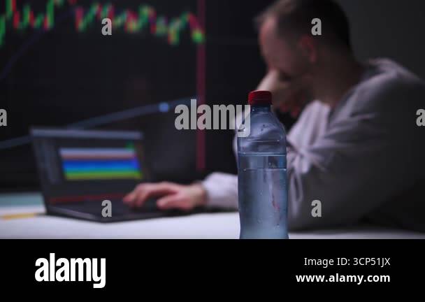 A man is sitting at a desk in an office, typing on a laptop in front of a large screen ...