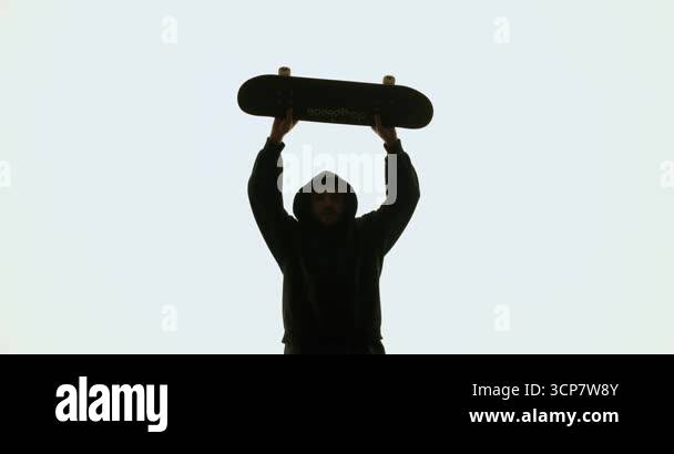 Silhouette of a young man in a hoodie holding a skateboard over his ...
