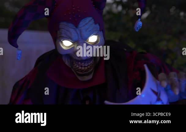 Scary Halloween jester clown at night with glowing eyes holding a white ...