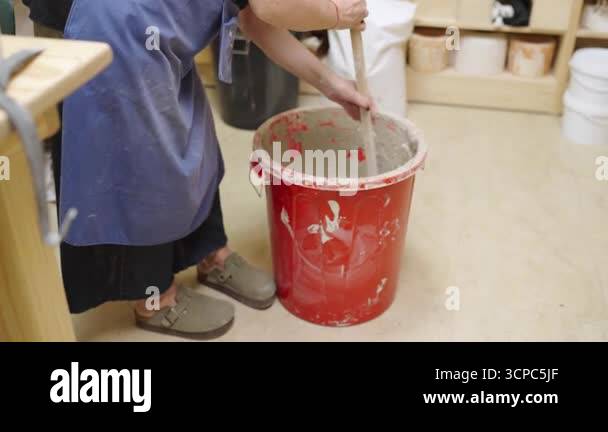 Professional potter mixing raw clay with water inside red bucket, using ...