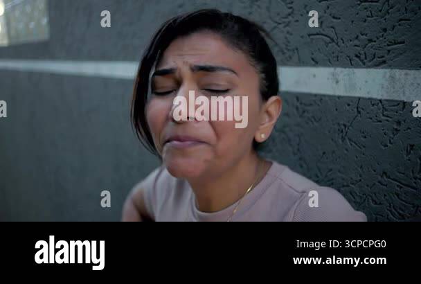 Latina woman hides face in hands while crying against wall, overwhelmed ...