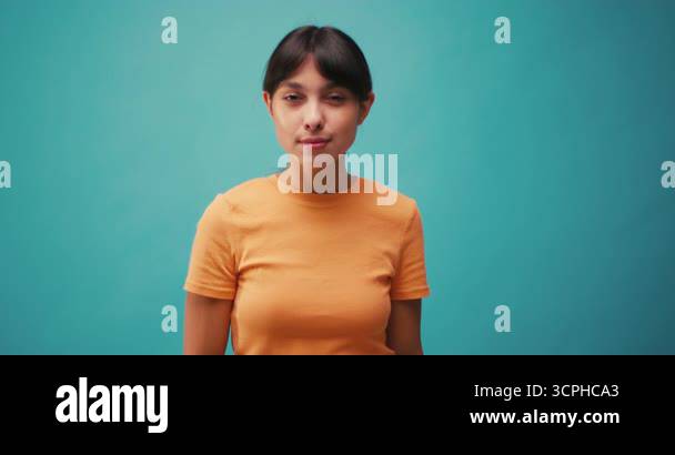 Young Hispanic woman in orange t-shirt looks curiously on blue backdrop ...