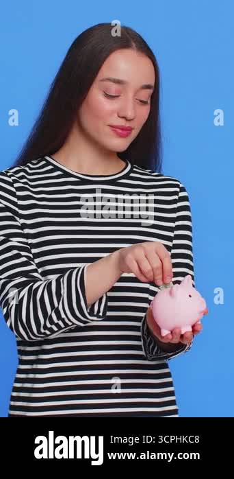 Happy rich young woman in striped blouse putting dollar pocket money cash into piggy bank saving ...