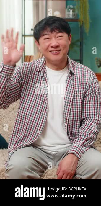 Asian man waves cheerful hello, raising hand with friendly smile ...
