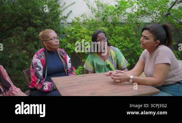 Four diverse women gather in backyard around wooden table, actively ...
