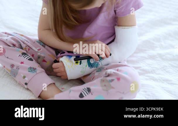 Adorable preschooler girl with a broken arm at home on the bed draws ...