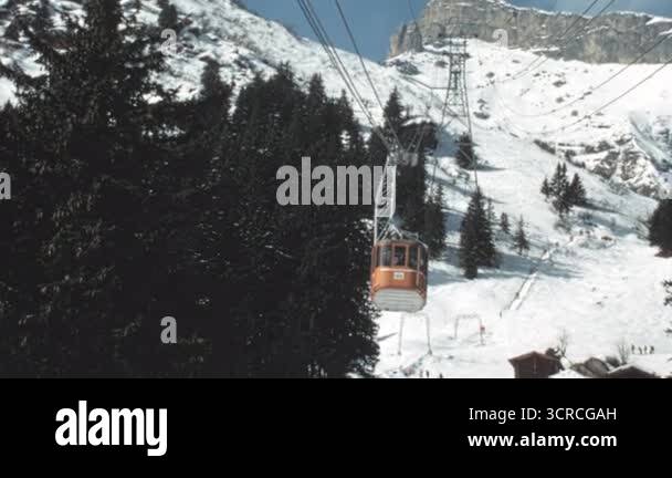 Orange and white cable car ascends a steep snowy mountain over an ...