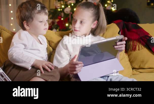 Brother and elder sister sit closely together on comfortable couch and ...