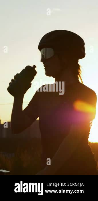 Woman cyclist is drinking water in park against sunset lights. Portrait ...