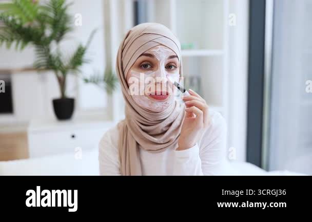 Smiling young Muslim woman wearing hijab applying facial mask with ...