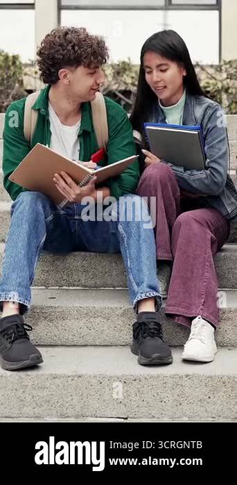Two university students are sitting on steps and sharing notes, helping ...
