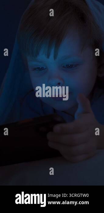 Little child lying under blanket at night, holding smartphone, playing ...