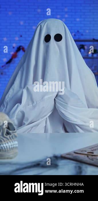 A person dressed as a ghost in a white sheet sits in a studio with blue ...