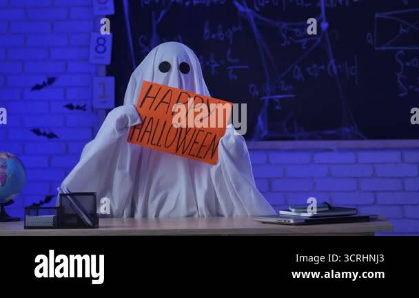A person dressed in a white ghost sheet holds a Happy Halloween sign in ...