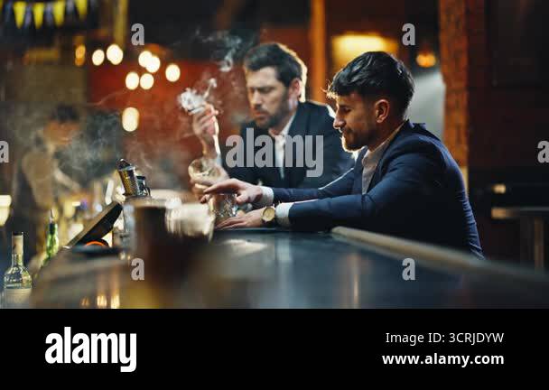 Two men drinking bar puffing smoke sitting in amber illuminated light ...