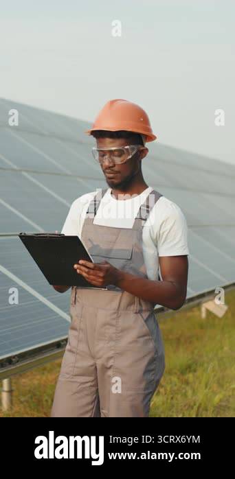 An African American technician in safety gear inspects solar panels ...