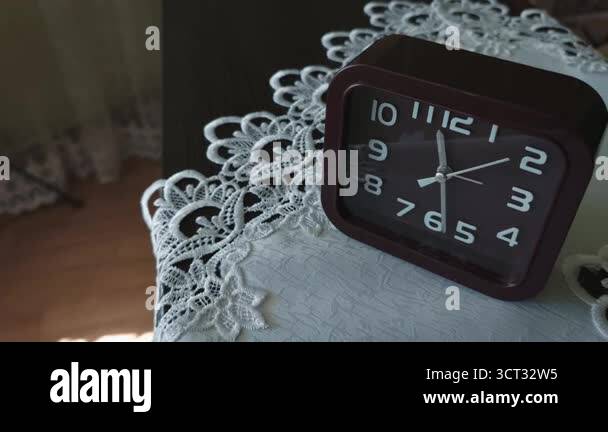 A square brown alarm clock rests on an elegant white lace doily on a bedside table. The camera ...