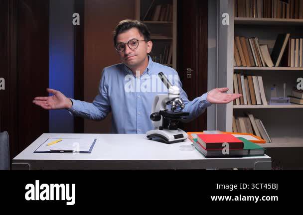 Puzzled scientist is shrugging his shoulders while sitting at a desk ...