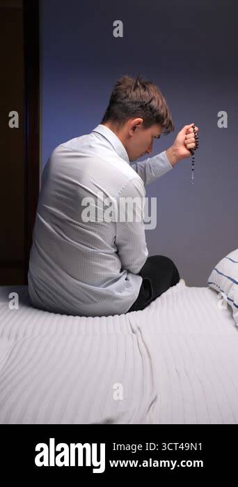 vertical video Man holding a rosary and praying on a bed in a subdued ...