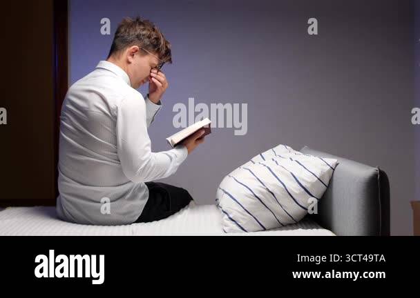 Man in glasses rubbing tired eyes while reading a book in bed, showing ...