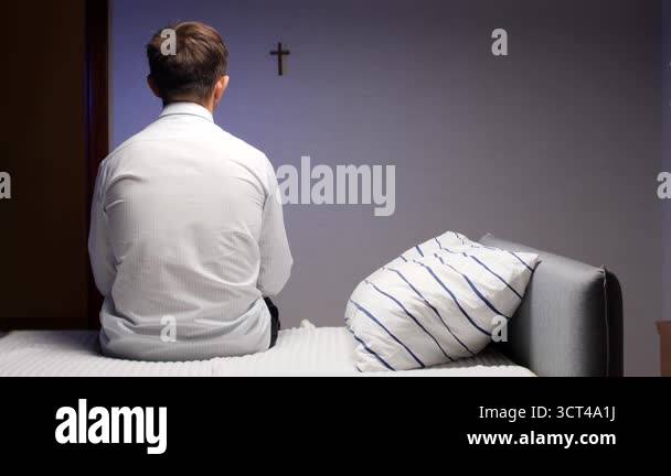 christian Man sits on a bed facing a wall cross, absorbed in quiet ...