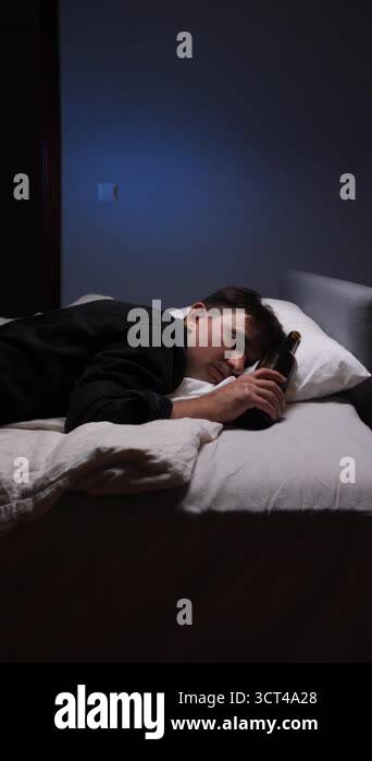 vertical video Drunk man sleeping on a bed, clutching a beer bottle in ...
