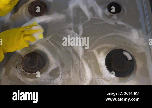 Hands in yellow gloves scrub a dirty gas stove with foam and sponge ...