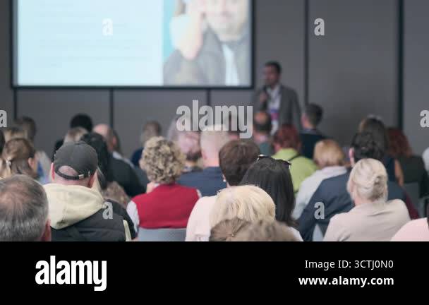 Crowd of professionals sits in rows facing large screen as speaker ...