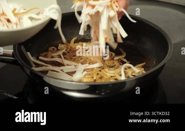 Female hands add sliced crab sticks to caramelized onion in a frying ...