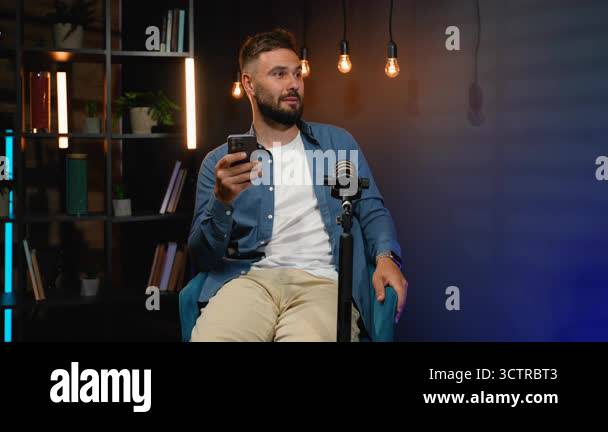 Young male influencer sitting in a modern studio, holding a smartphone ...