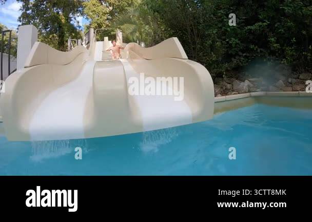 Teen boy sliding down curved water slide into pool captured in slow ...