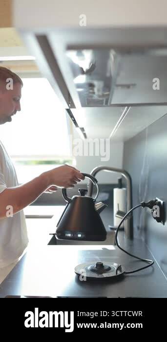 A man is standing in a bright modern kitchen, filling an electric ...