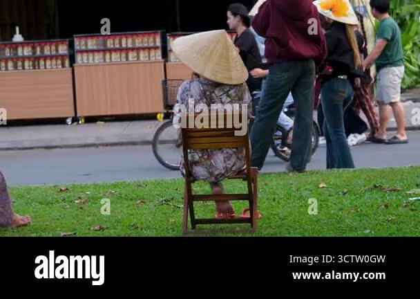 Women in Vietnamese hats sitting outdoors on chairs observing tourists ...