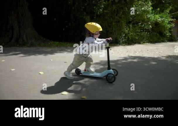Toddler falling off scooter on park path wearing yellow helmet, playful ...