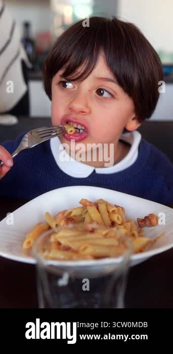 Boy eating pasta with fork at dining table, leaning forward and opening ...