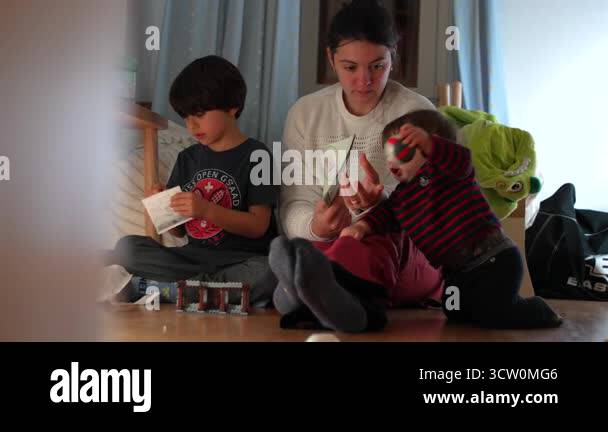 Mother seated with children in evening at home, toddler reaching while ...