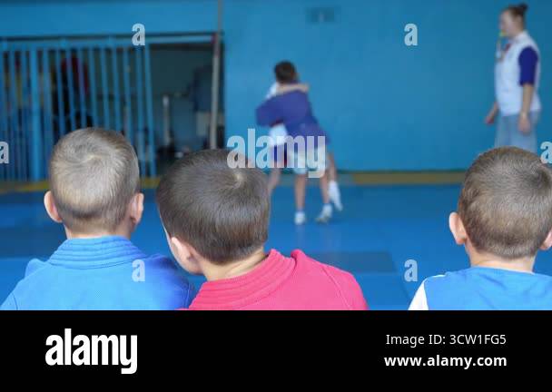 Three young boys in sambo uniforms watch a sports match on the mat ...
