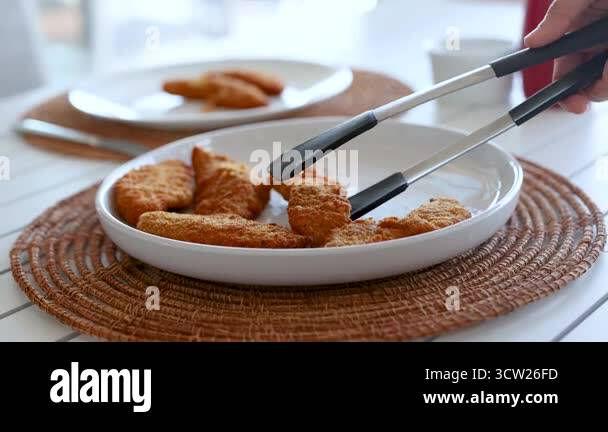 Tongs place crispy fried chicken pieces onto a white plate on a woven ...